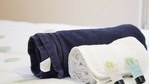Toalha azul-marinho dobrada com cuidado e uma toalha branca, com duas embalagens de Shampoo e Condicionador Revitalizante sobre uma cama com lençol branco e desenhos de folhas verdes.
