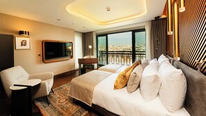 Quarto de hotel moderno com uma cama grande, área de estar com uma cadeira e mesa, televisão presa à parede e varanda com vista para a cidade.