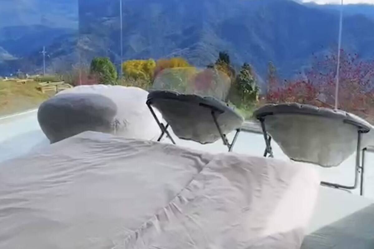 Uma cama com colchão branco e estrutura de metal, janela com vista para montanhas e árvores, e piso de madeira.