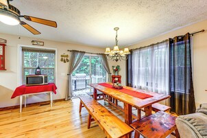 Uma área de refeições com mesa de madeira, bancos, ventilador de teto e vista para o exterior através de grandes janelas.