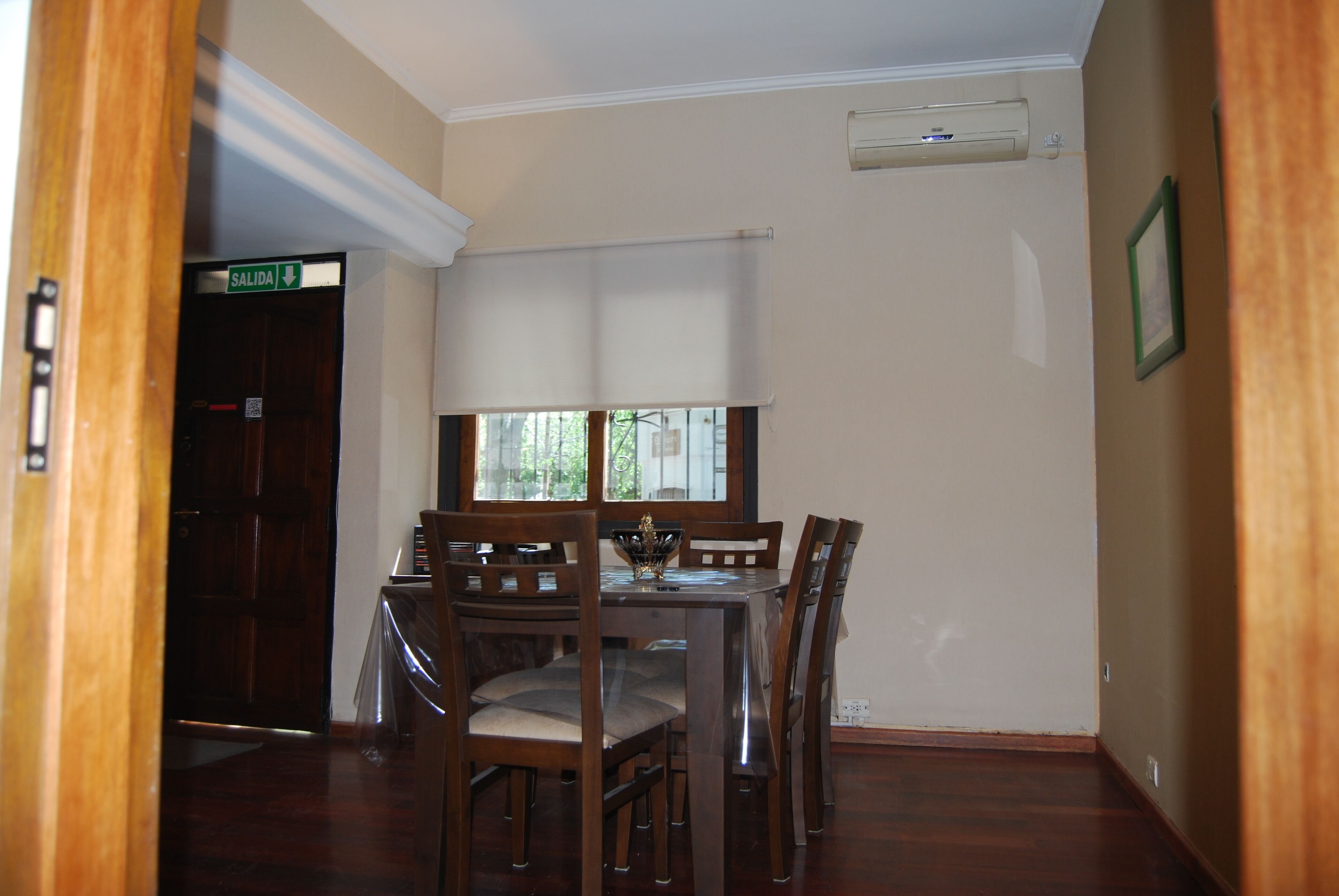 Uma área de refeições com mesa e cadeiras, uma porta com a placa 'SALIDA' e uma janela com persianas.