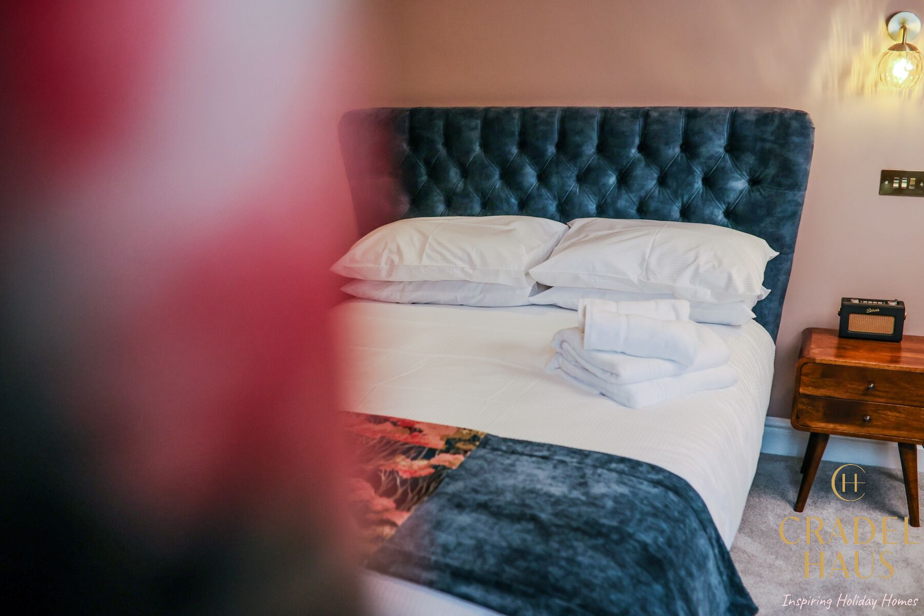 Cama bem arrumada com lençóis brancos e cabeceira azul escura. Um criado-mudo de madeira com um rádio fica ao lado da cama.