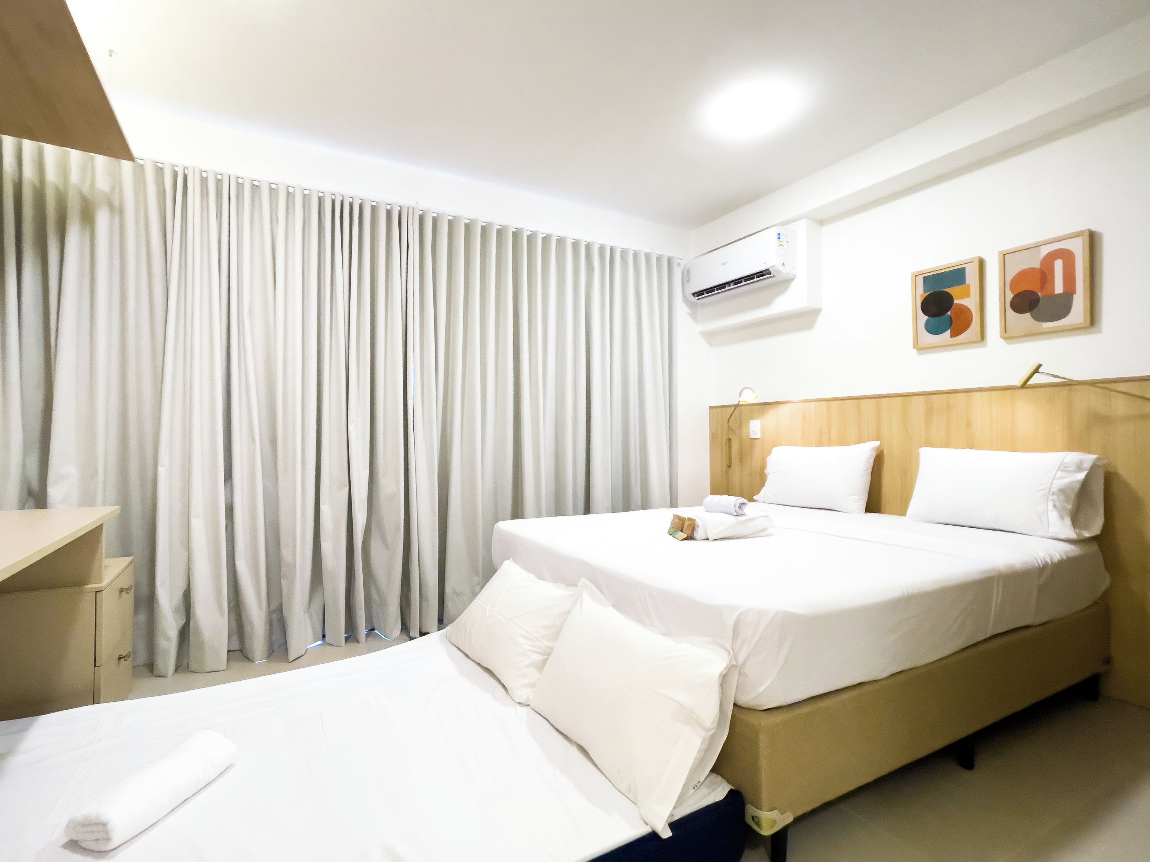 Quarto de hotel com cama, escrivaninha, cadeira, janela com cortinas, aparelho de ar condicionado e duas obras de arte fixadas na parede.