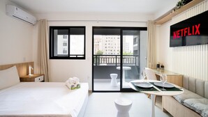 Quarto de hotel com cama, mesa de jantar com cadeiras, sofá, televisão e varanda com vista para edifícios.