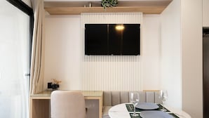 Uma sala de jantar moderna com uma mesa de madeira, um banco acolchoado e uma mesa redonda posta para dois. Uma televisão de tela plana está montada acima do banco, e há uma pequena planta na parede.