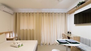 Quarto de hotel com cama, mesa de cabeceira com abajur, pequena mesa com um jogo americano verde e duas taças de vinho, sofá e televisão fixada na parede.