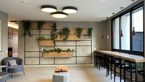 Um interior moderno com uma longa mesa de madeira, várias cadeiras, um banquinho de madeira redondo e uma área de estar com um sofá e poltrona cinzas.