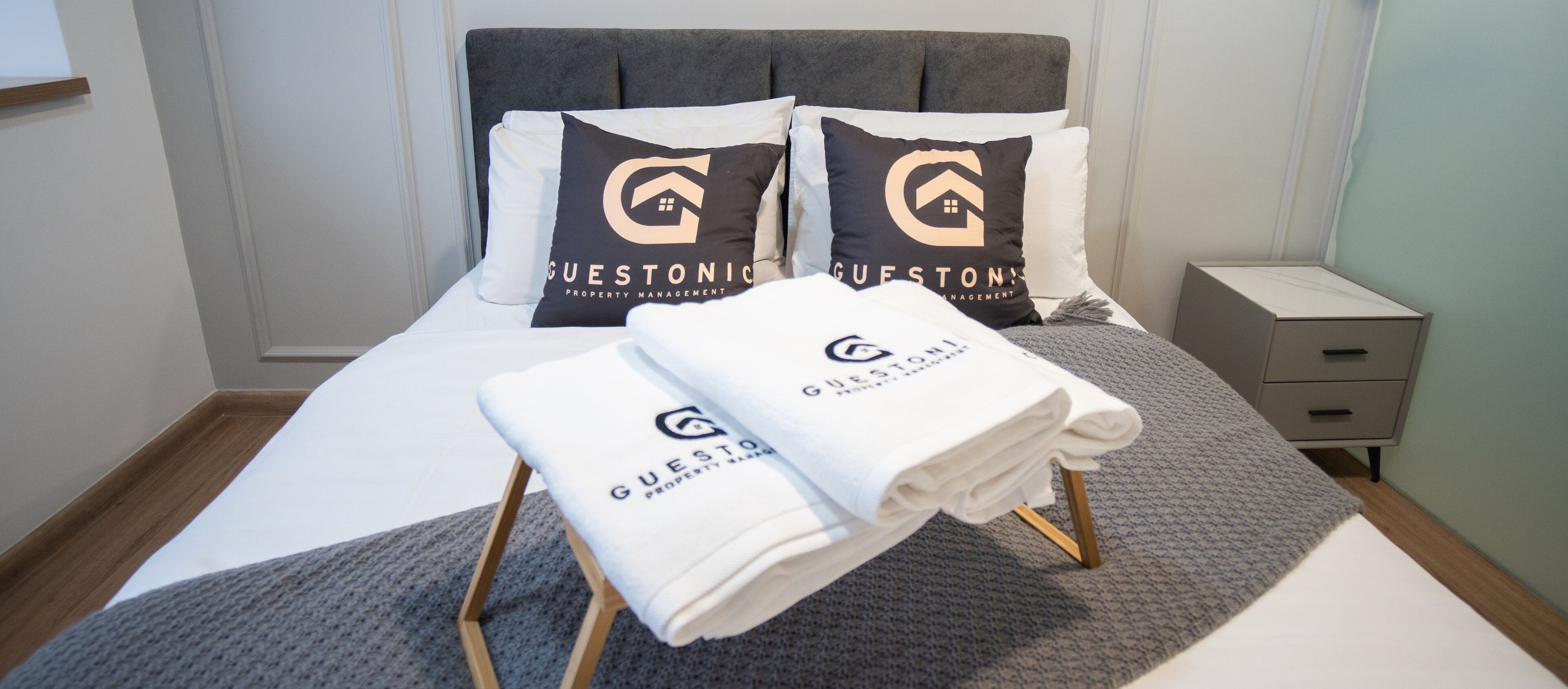 Cama bem arrumada com duas almofadas e duas toalhas dobradas, cada uma com um logotipo.