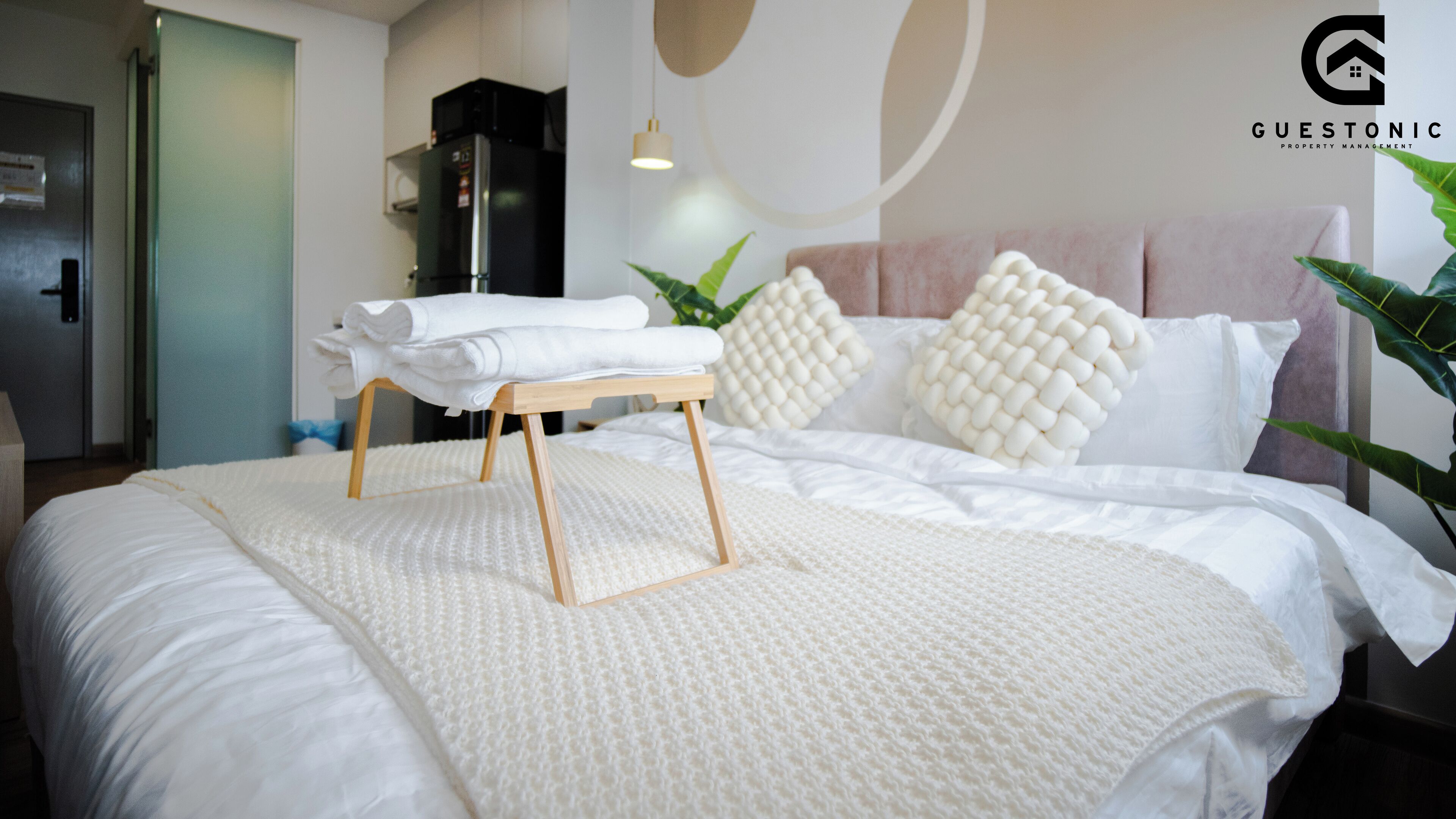Uma cama bem arrumada com edredom branco e travesseiros, um banquinho de madeira com toalhas dobradas, uma luminária moderna e uma planta.