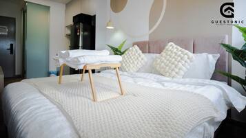 Uma cama bem arrumada com edredom branco e travesseiros, um banquinho de madeira com toalhas dobradas, uma luminária moderna e uma planta.