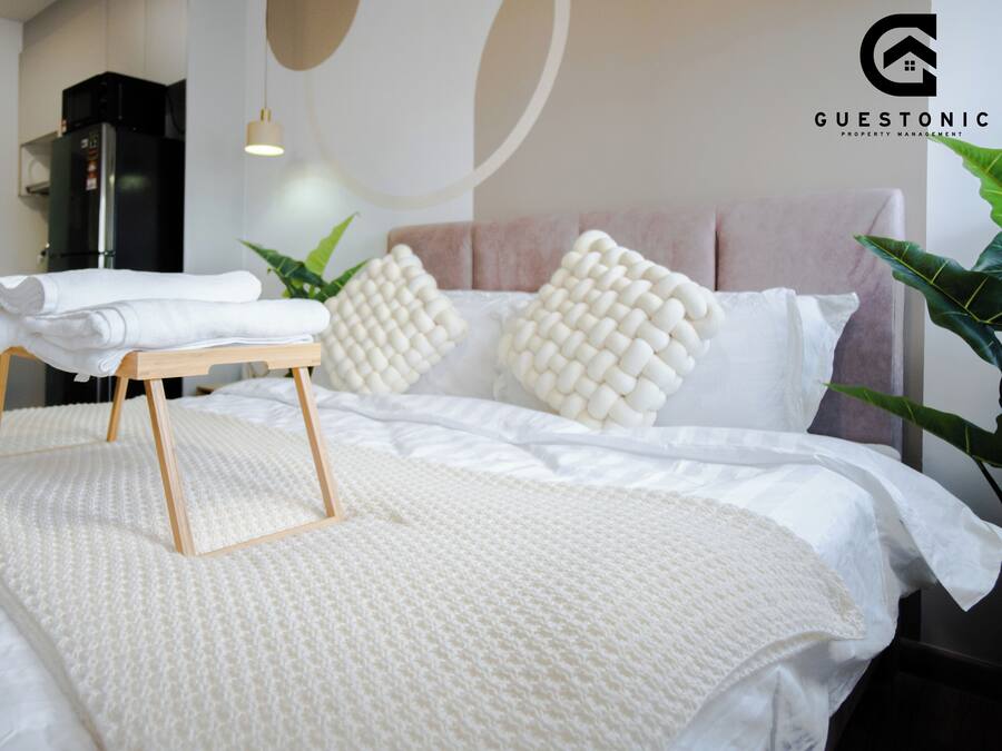 Uma cama bem arrumada com edredom branco e travesseiros, um banquinho de madeira com toalhas dobradas, uma luminária moderna e uma planta.