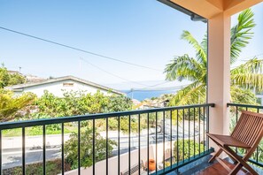Uma varanda com cadeira de madeira, guarda-corpo e vista para a rua e vegetação.