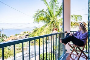 Varanda com vista para o mar, palmeiras e uma pessoa sentada numa cadeira.
