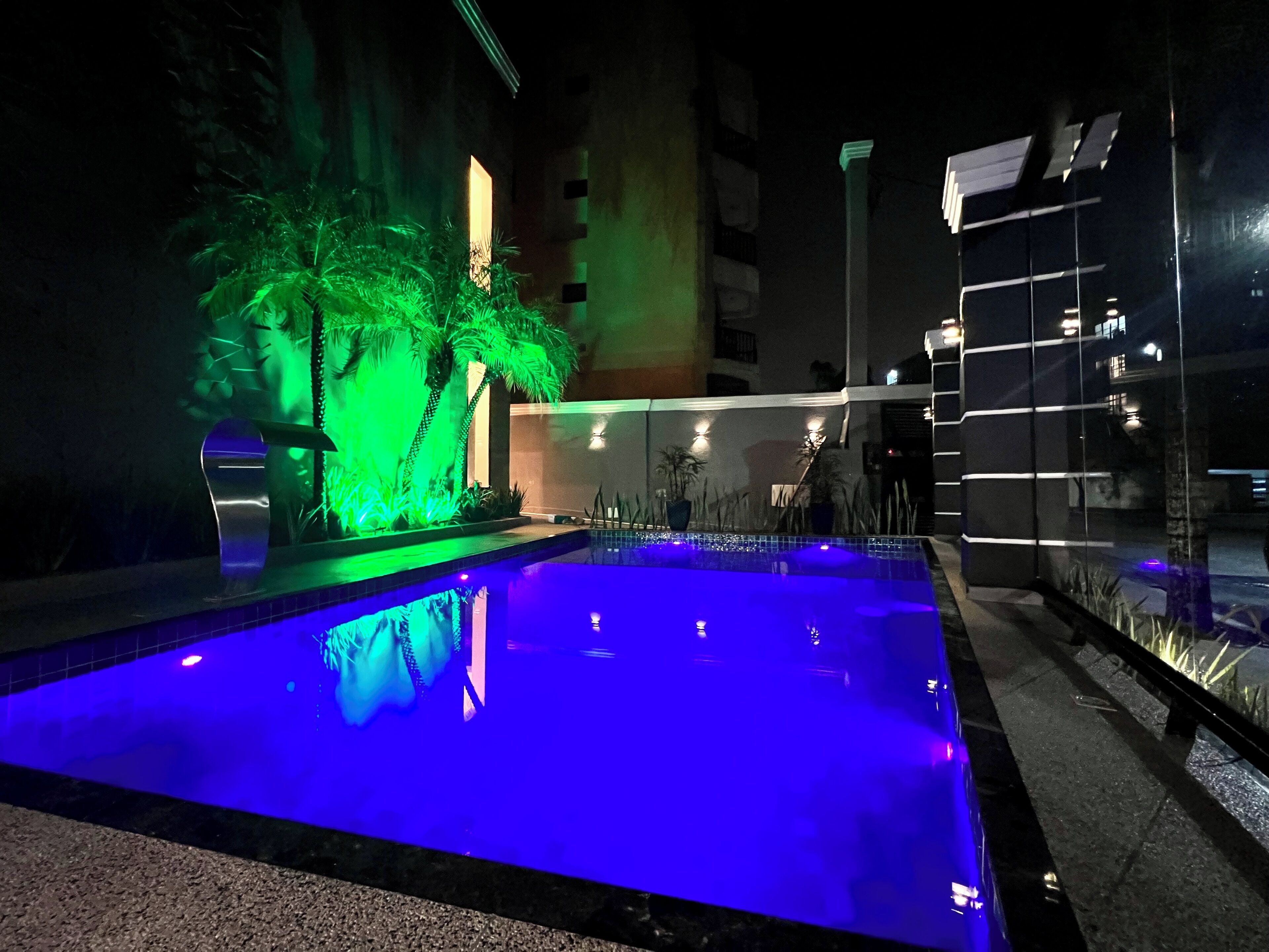 Piscina noturna iluminada em verde e azul, cercada por edifícios e palmeiras.