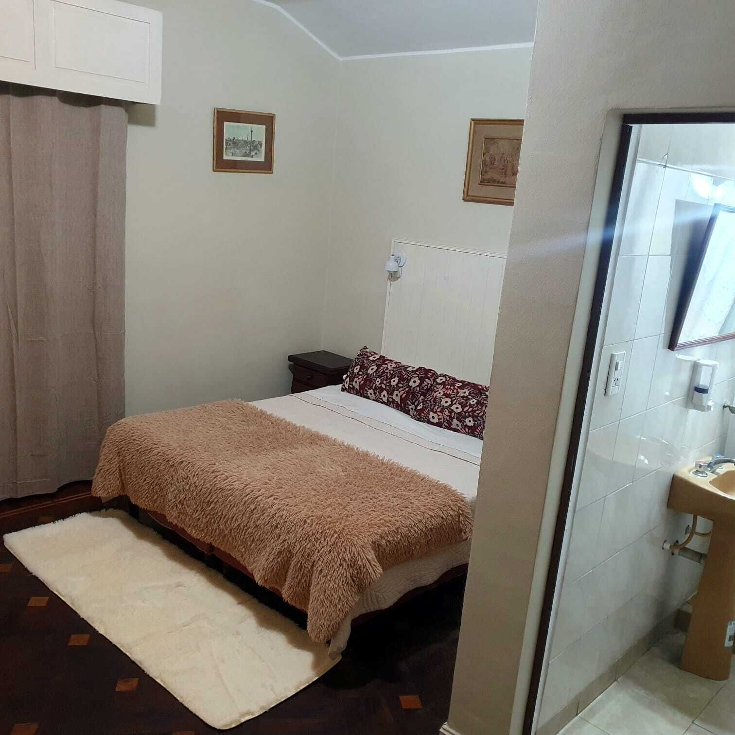 Quarto com cama, mesa de cabeceira, janela com cortinas, quadro na parede e banheiro com pia e espelho.