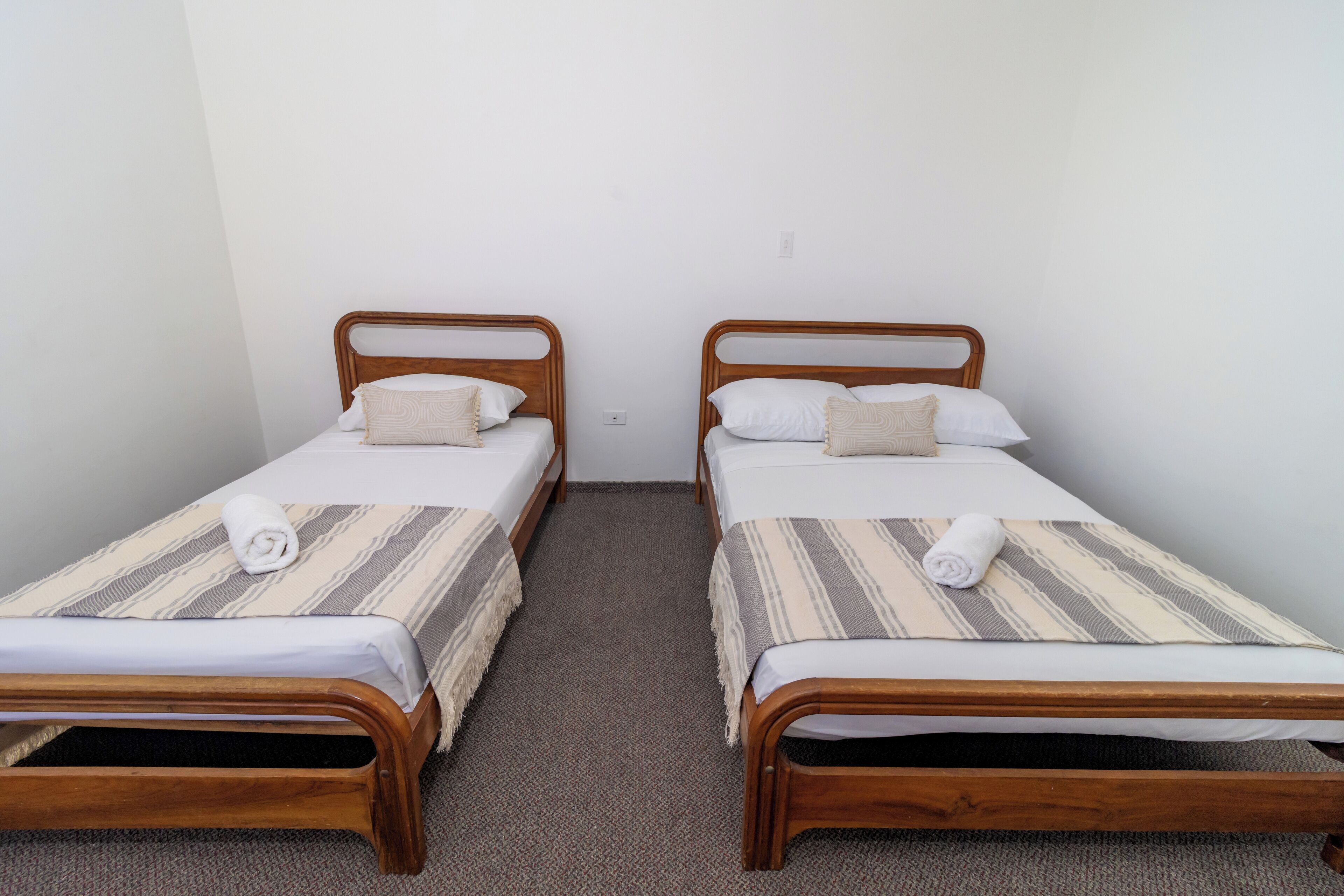 Duas camas de solteiro com estrutura de madeira e roupa de cama listrada, um piso coberto por carpete e uma parede branca sem adornos.