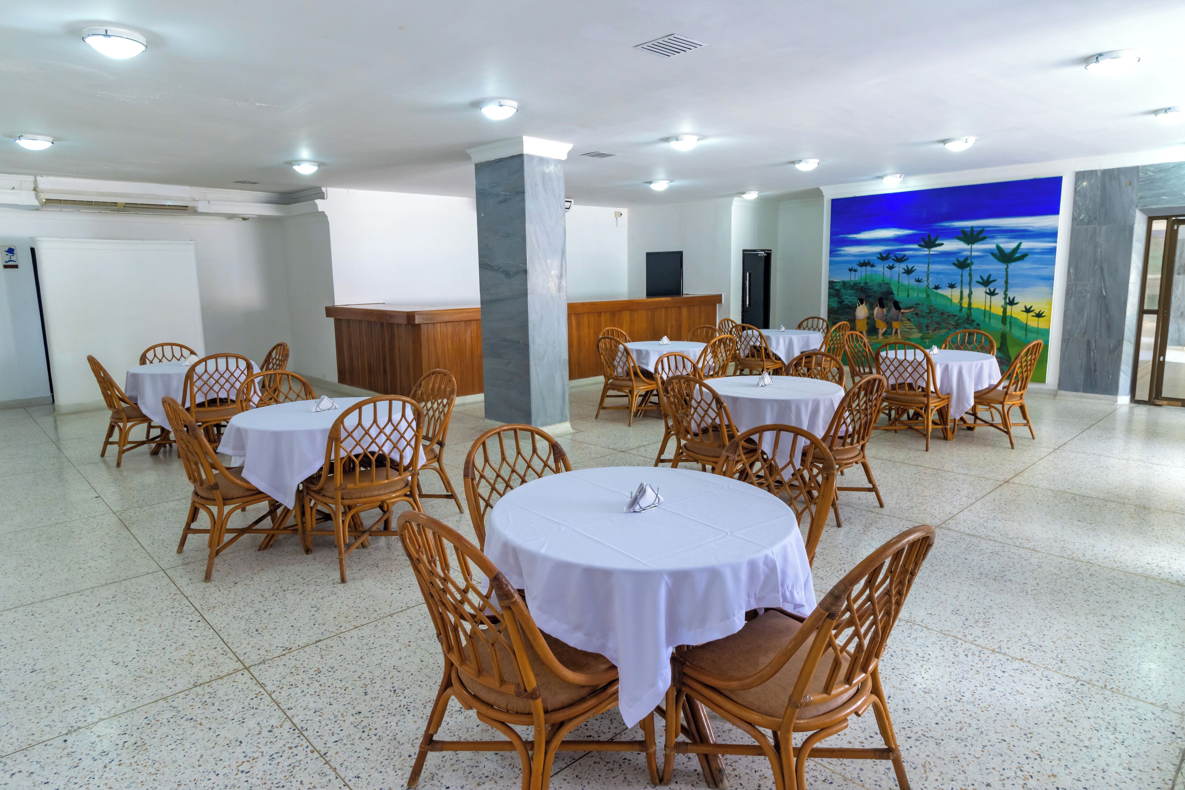Uma sala de jantar com mesas redondas cobertas por toalhas brancas, cadeiras de madeira e um mural na parede.