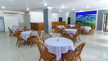 Uma sala de jantar com mesas redondas cobertas por toalhas brancas, cadeiras de madeira e um mural na parede.