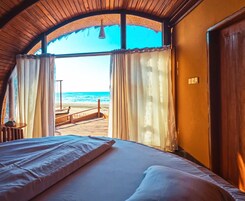 Quarto com teto de madeira, teto curvo e uma grande janela com cortinas, com vista para uma praia.