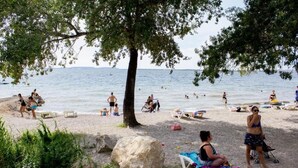 Uma praia com pessoas se banhando, nadando e caminhando. Há cadeiras de praia e guarda-sóis na areia.