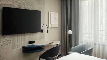 Quarto de hotel moderno com televisão de tela plana na parede, uma mesa redonda com cadeira, mesa de cabeceira e uma janela grande com cortinas.