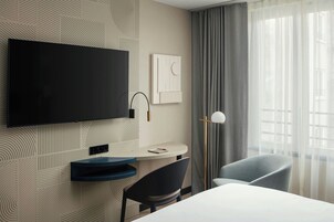 Quarto de hotel moderno com televisão de tela plana na parede, uma mesa redonda com cadeira, mesa de cabeceira e uma janela grande com cortinas.