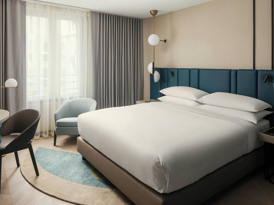 Quarto de hotel com uma cama grande, uma escrivaninha com cadeira, luminária e janela com cortinas.