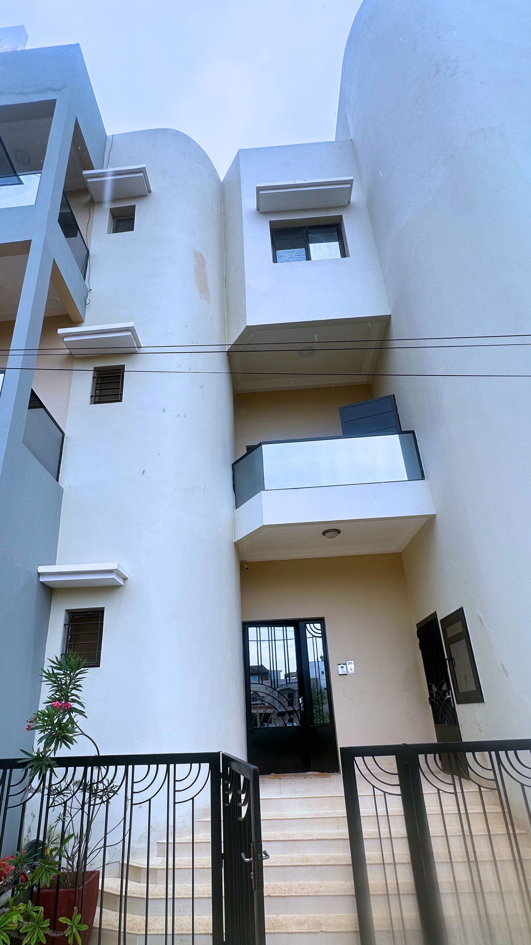 Um edifício residencial de vários andares com varandas, entrada com portão e escadaria que dá acesso ao prédio.