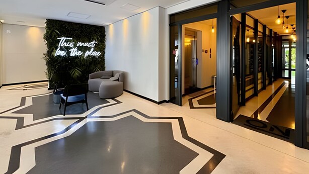 Um lobby moderno com design geométrico no chão, uma parede verde com um letreiro de neon e uma área de estar.