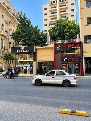 Vista da rua com um carro branco, um prédio com a placa 'Valencia Burger' e uma loja 'Shalan'.