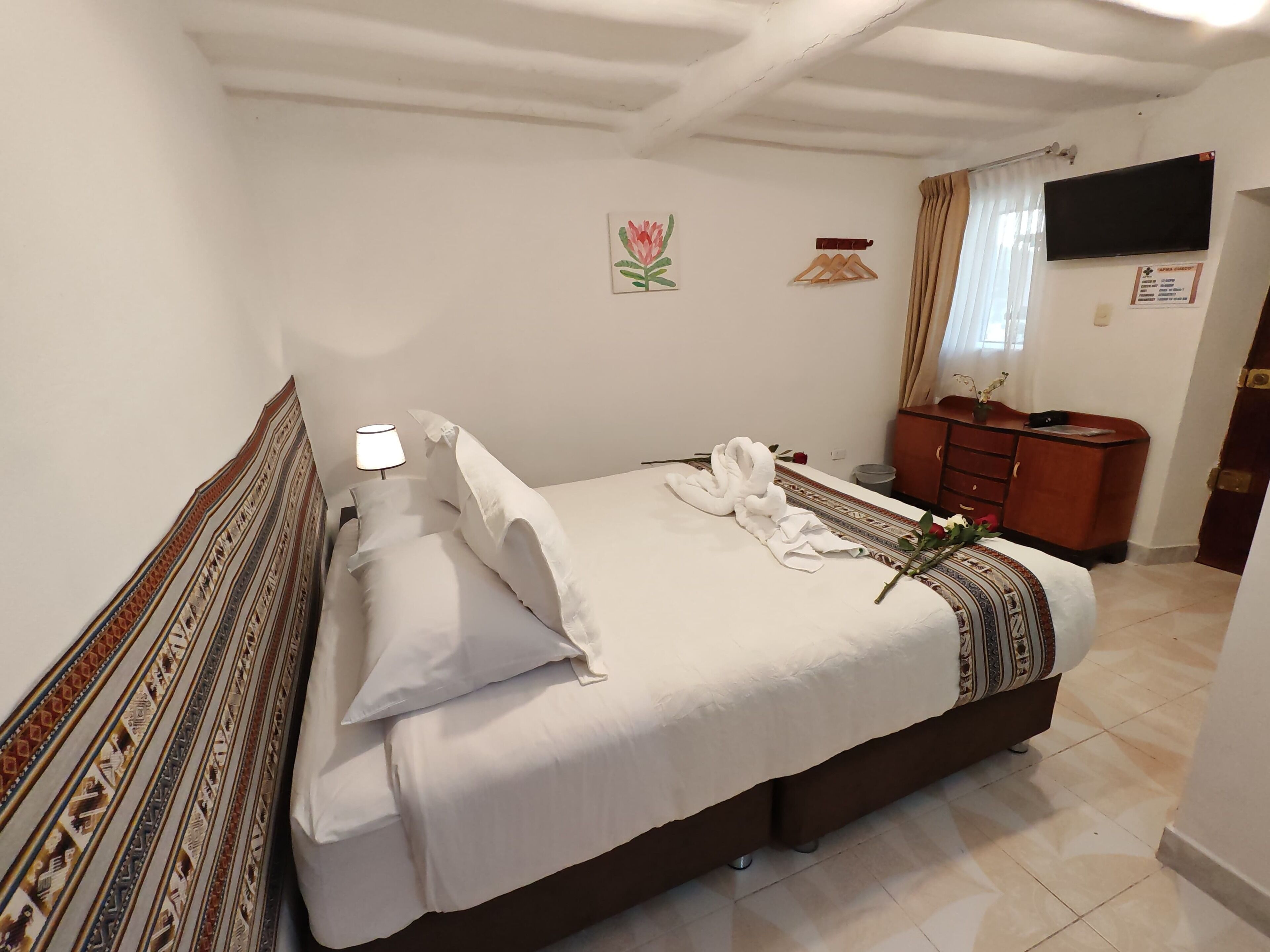 Uma cama bem arrumada com toalha e decoração de rosa, um aparador de madeira e uma televisão na parede.