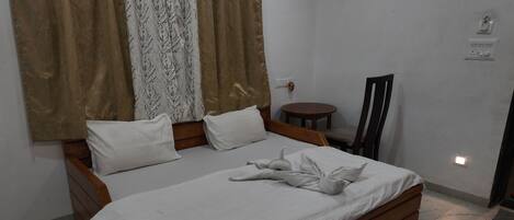 Cama bem arrumada com lençóis brancos e toalha, cabeceira de madeira, mesa redonda pequena e uma cadeira em um quarto simples com paredes brancas e piso de mármore.
