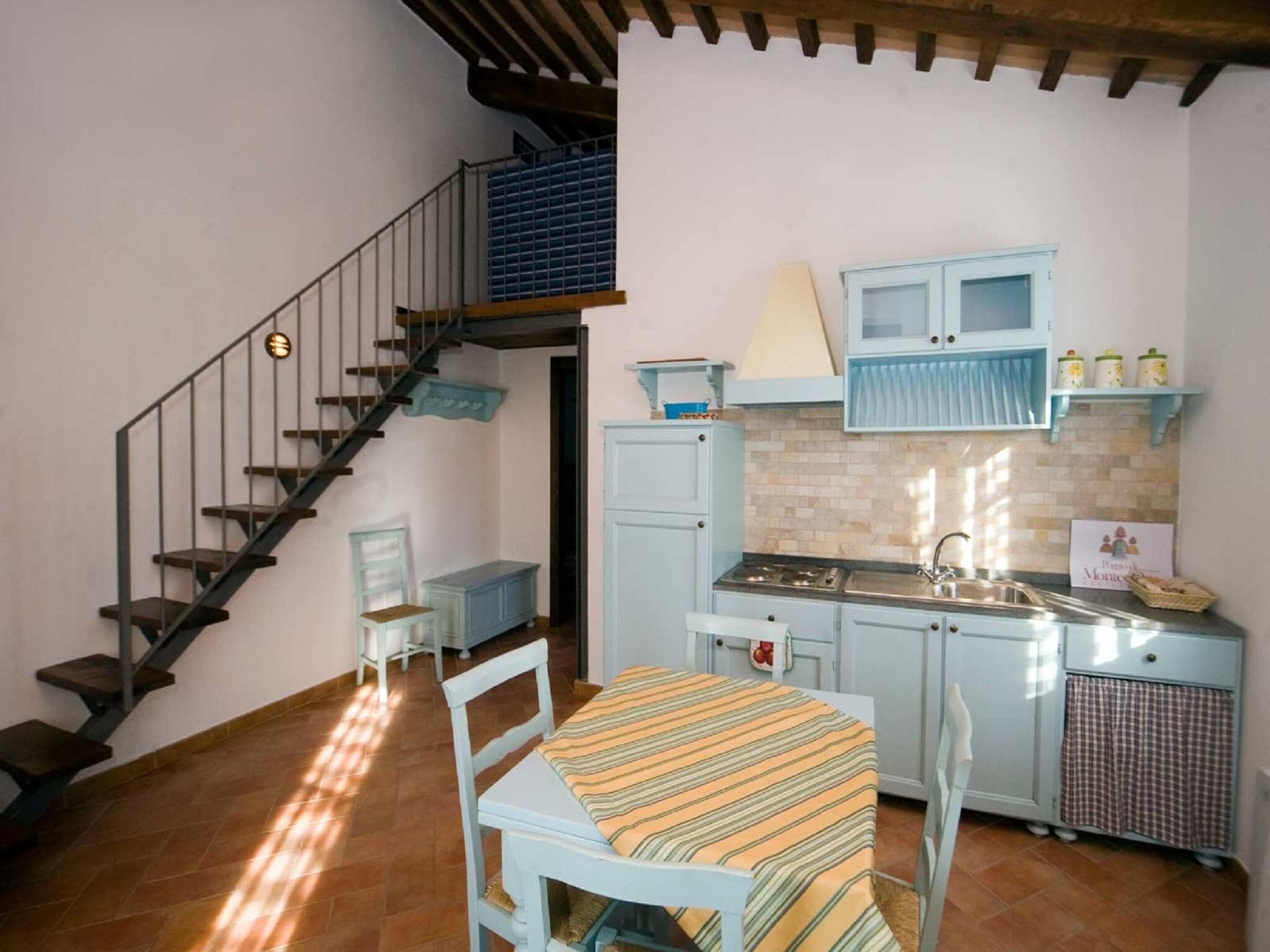 Uma cozinha compacta com painel de azulejos, fogão e pia. Há uma mesa de jantar com cadeiras, uma escada com guarda-corpo e uma pequena janela.
