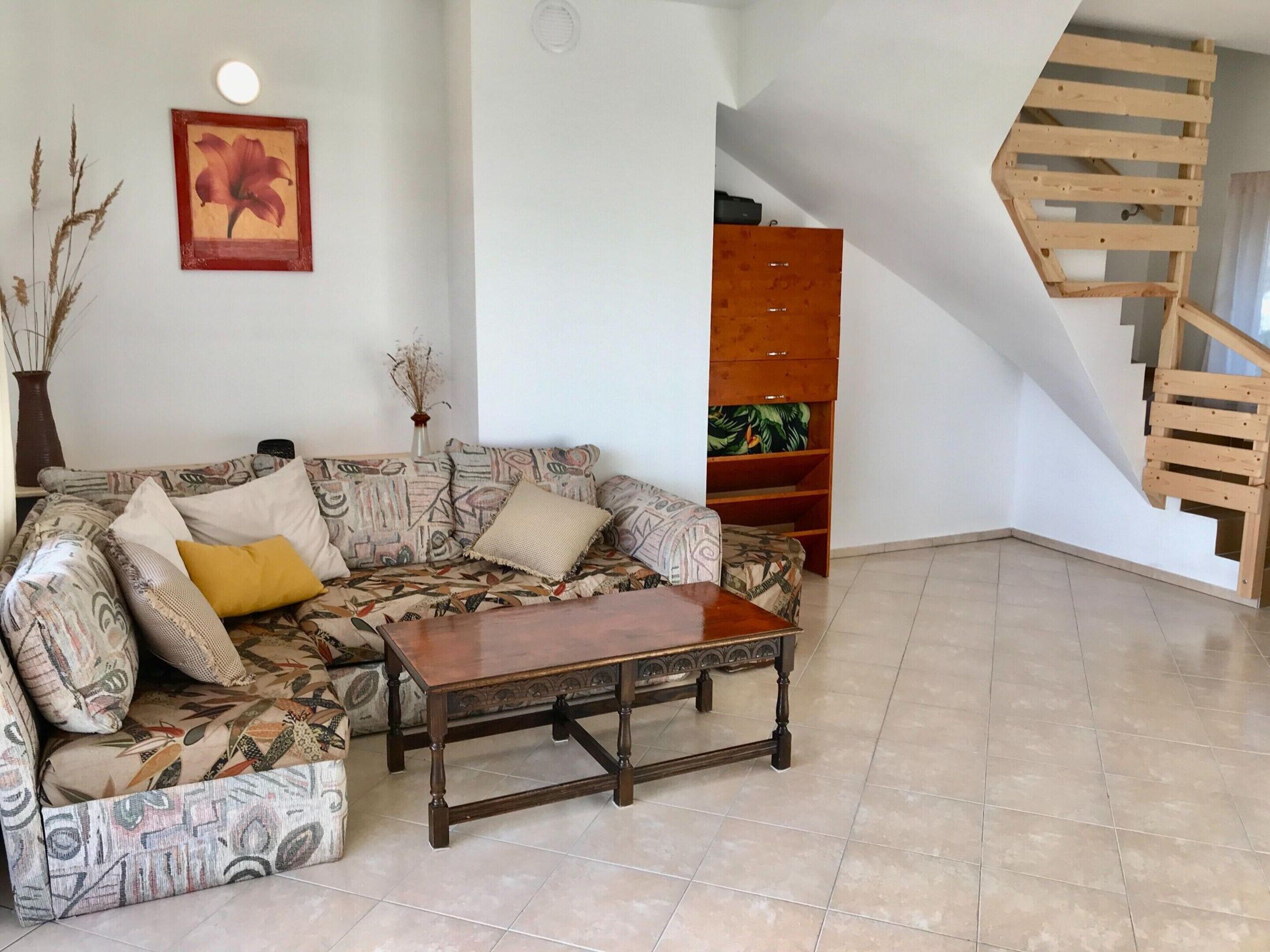 Uma sala de estar com um sofá estampado, uma mesa de centro de madeira, uma escada com corrimão de madeira e um luminário fixado na parede.