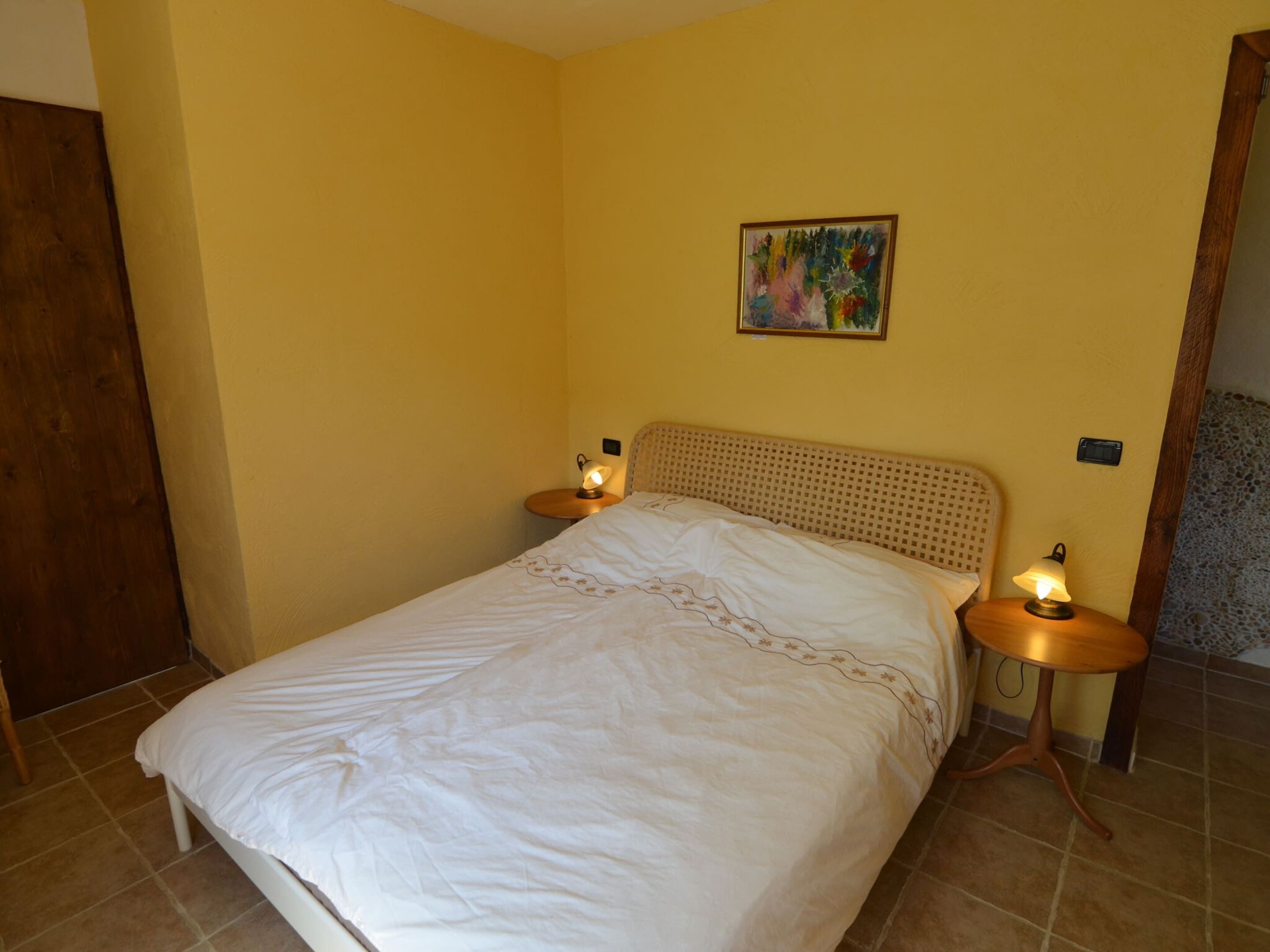 Quarto com cama, duas mesas de cabeceira com luminárias, um quadro na parede e porta de madeira.