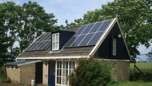 Uma casa com telhado inclinado, painéis solares, fachada de tijolos e um pequeno alpendre frontal.