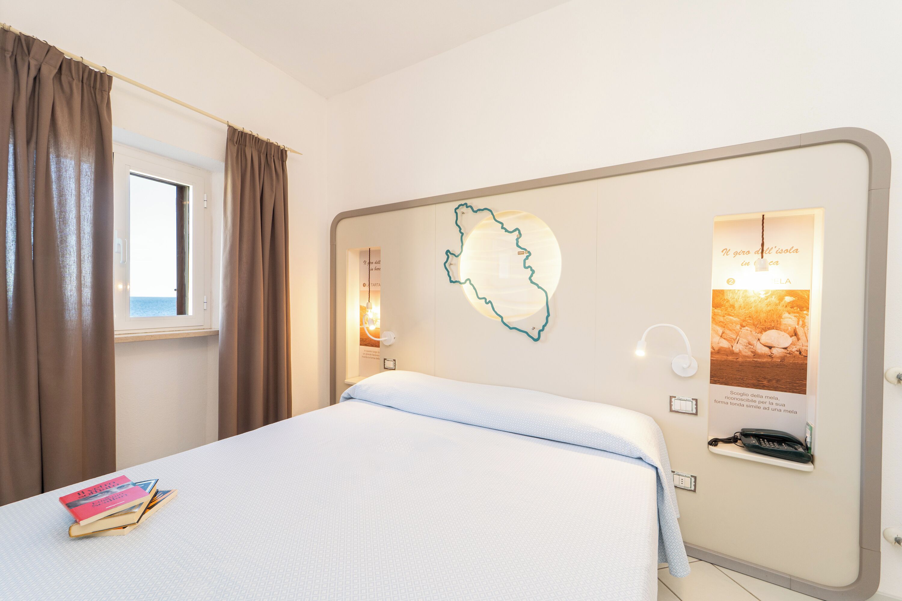 Quarto de hotel com cama, janela, cortinas, luminária na parede, uma obra de arte na parede e uma prateleira pequena com um telefone.