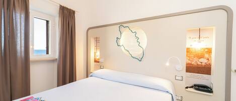 Quarto de hotel com cama, janela, cortinas, luminária na parede, uma obra de arte na parede e uma prateleira pequena com um telefone.
