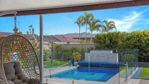 Uma área moderna de piscina ao ar livre com uma cadeira suspensa, paredes de vidro transparente e vista para o bairro.