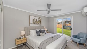 Quarto com cama, mesa de cabeceira, cadeira, ventilador de teto e uma janela grande com vista para uma casa e jardim.