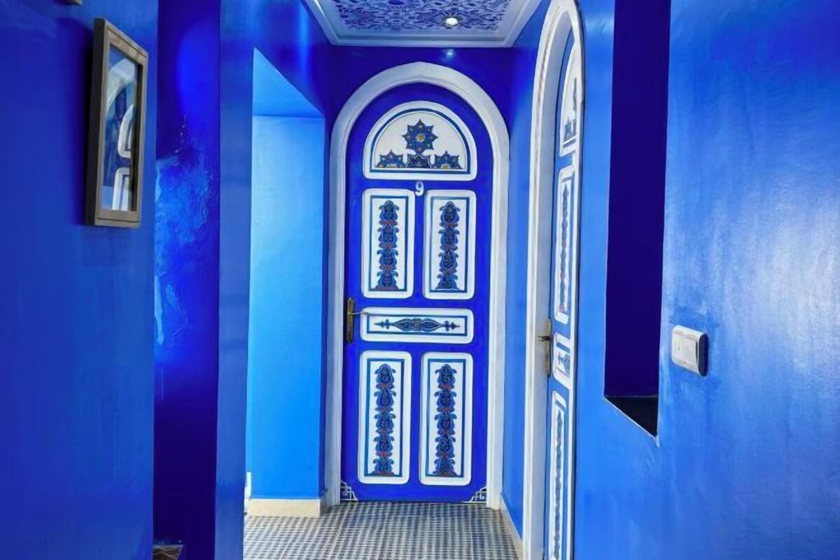 Corredor com porta e paredes azuis, que dá acesso a um quarto com piso estampado.