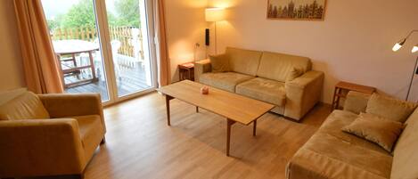 Sala de estar com sofá, poltrona e mesa de centro. Há uma porta de vidro deslizante que dá para uma área externa com mesa e cadeiras. Um abajur e um quadro estão na parede.
