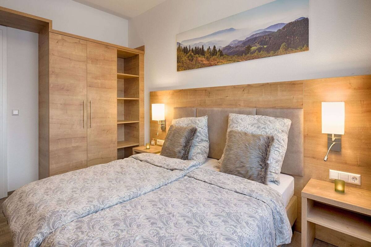 Quarto com cama, cabeceira de madeira, abajures e pintura de uma paisagem montanhosa.