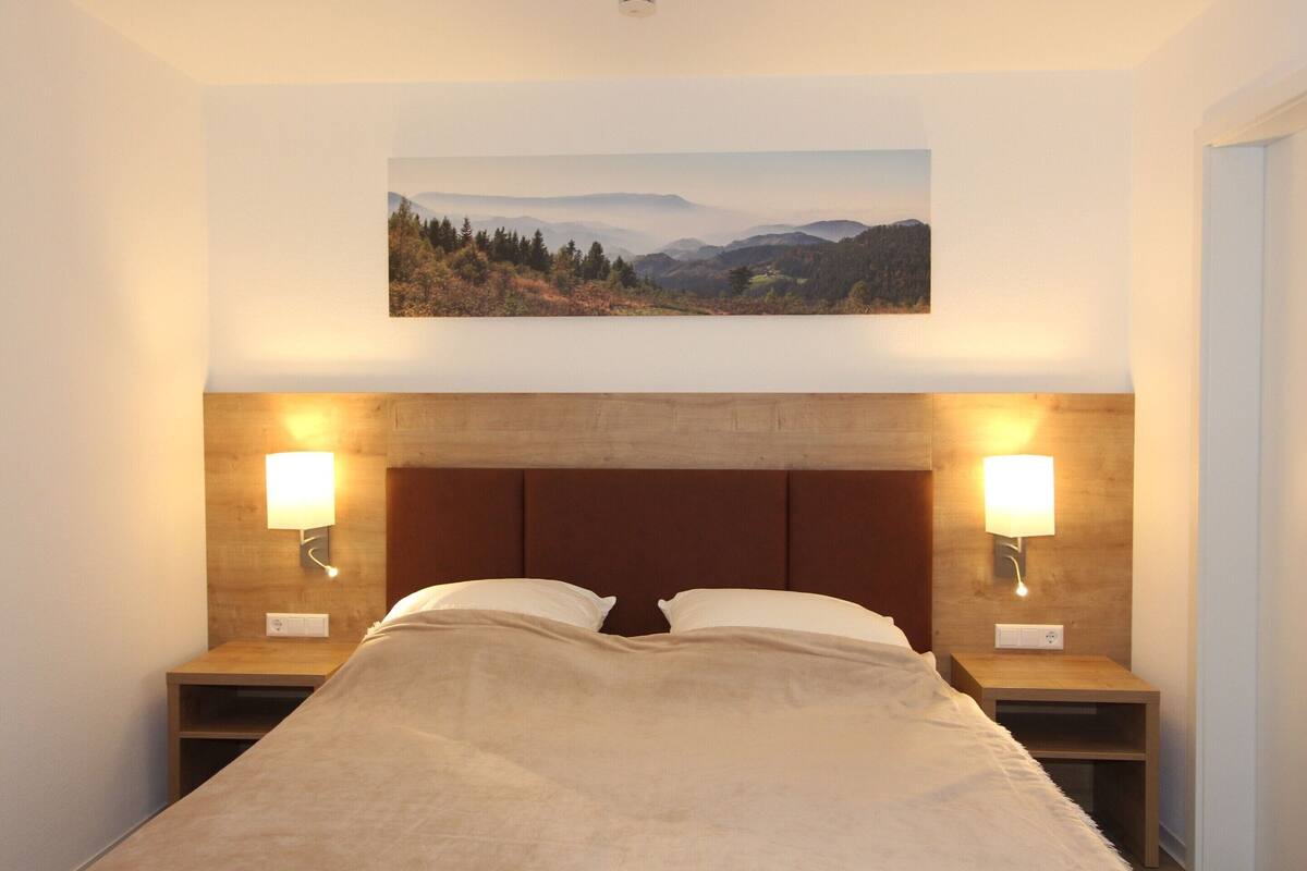 Uma cama bem arrumada com cabeceira, duas mesinhas de cabeceira com abajures, e um quadro de paisagem acima da cabeceira.