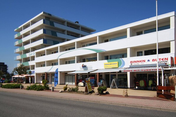 Um edifício de vários andares com fachada branca, varandas e lojas, incluindo um centro de informações turísticas e uma loja de roupas.