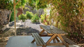 Mesa e banco de madeira sob uma pérgula em um jardim com diversas plantas e uma parede de pedra.