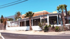 Um edifício com arquitetura moderna, com teto plano e grandes janelas. Palmeiras e um céu azul claro ao fundo.