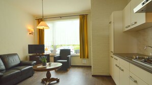 Sala de estar com sofá, mesa de centro e televisão. Há uma janela com cortinas e vista para o exterior. A cozinha possui armários e pia.
