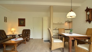 Sala de estar com sofá, duas poltronas, mesa de centro e mesa de jantar com cadeiras. Há uma área de cozinha com armários e luminária pendente.