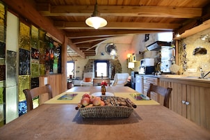 Uma mesa de jantar de madeira com uma cesta de palha contendo maçãs e nozes, um vaso com flores, e vista para uma cozinha e sala de estar com paredes de pedra e vigas de madeira.